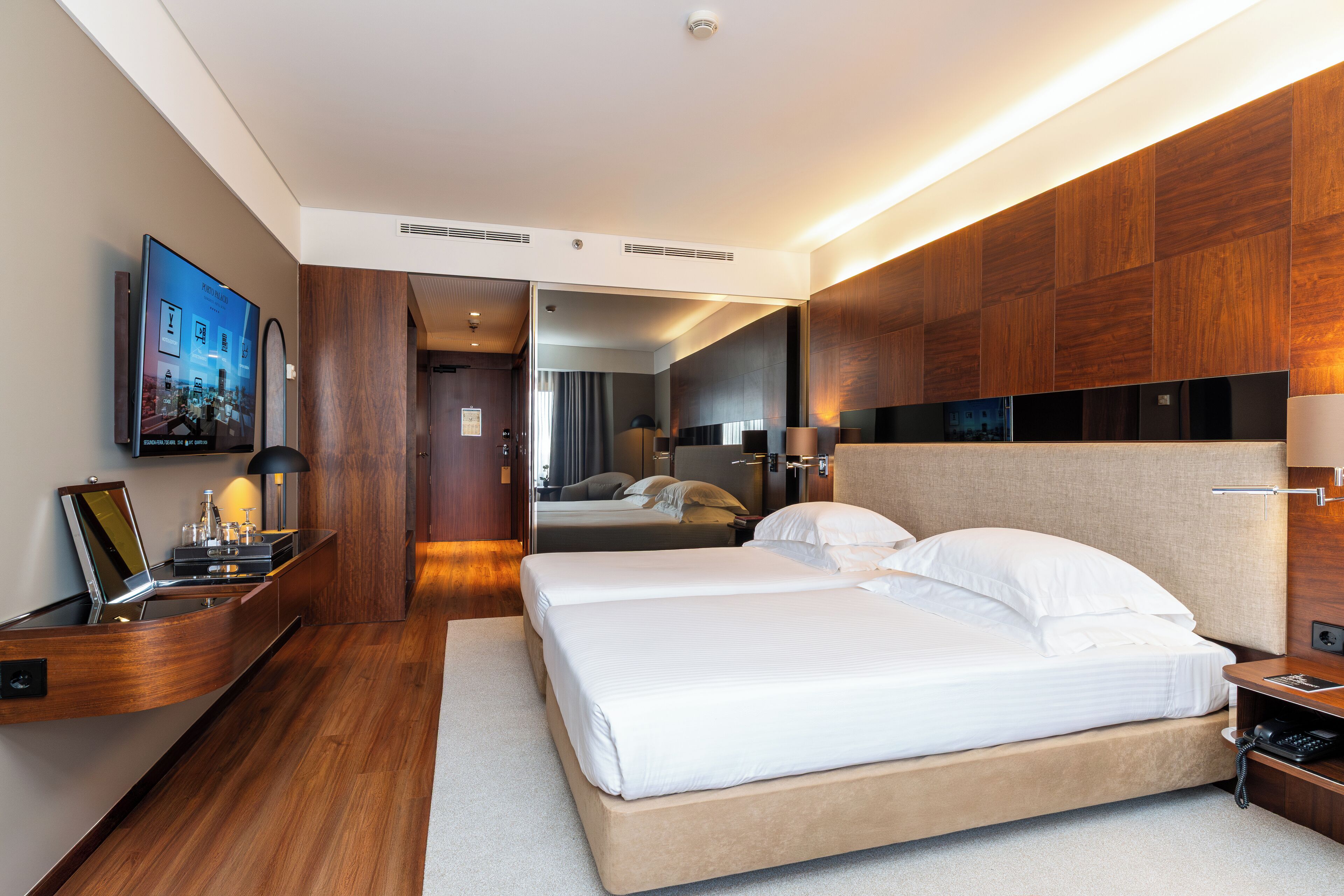 Quarto de hotel moderno com uma cama grande, uma escrivaninha com computador, televisão de tela plana e um banheiro visível por uma porta aberta.