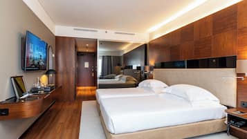 Quarto de hotel moderno com uma cama grande, uma escrivaninha com computador, televisão de tela plana e um banheiro visível por uma porta aberta.