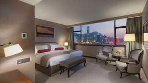 Quarto de hotel com uma cama grande, uma escrivaninha, duas poltronas e vista para a paisagem urbana.