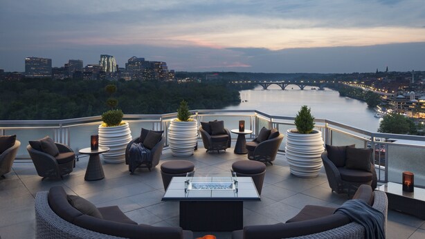 Terraço no telhado com área de estar ao ar livre, mesa com tampo de vidro e vista para a paisagem urbana e rio ao anoitecer.