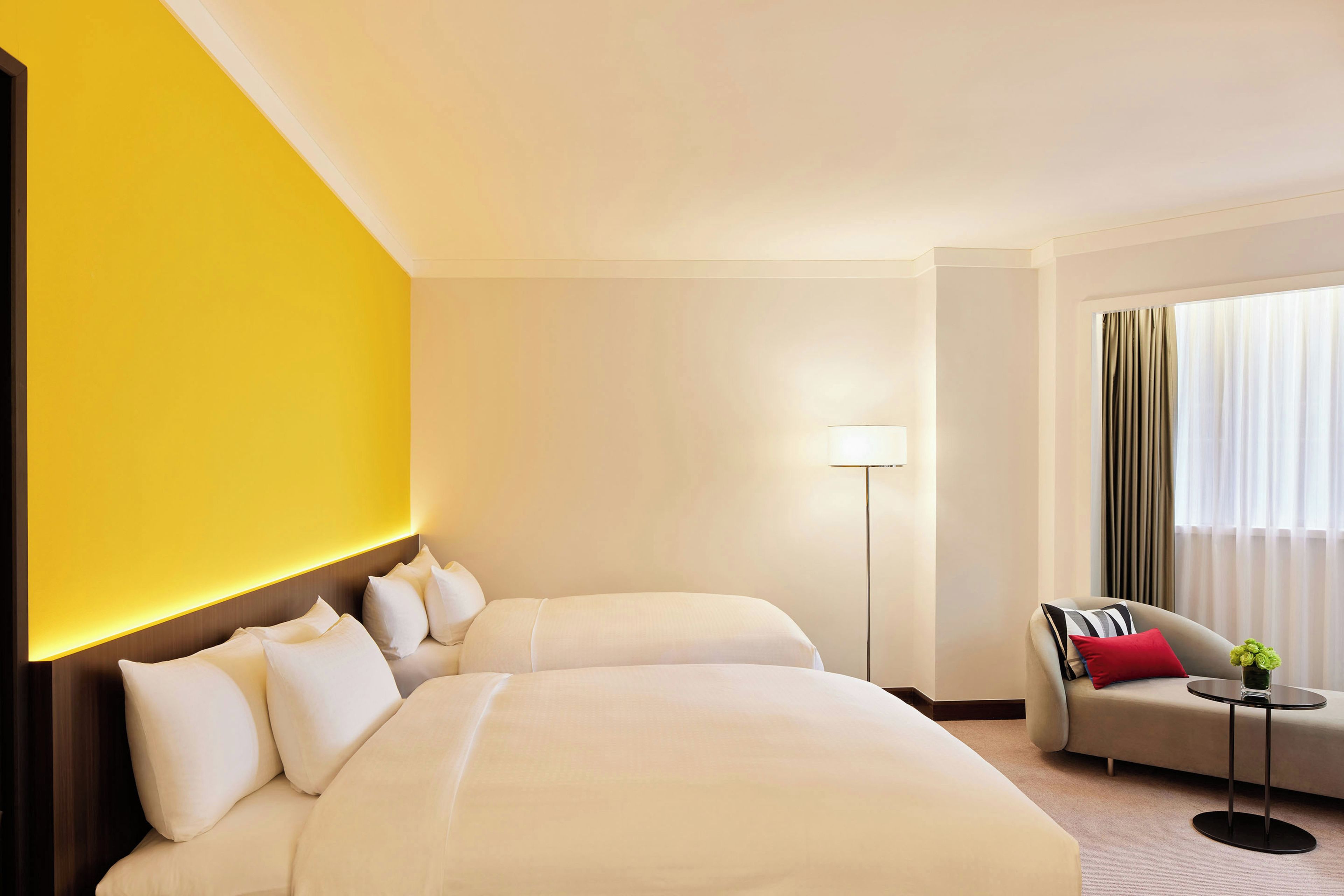 Quarto de hotel com duas camas, um sofá, uma luminária e uma pequena mesa com um vaso.