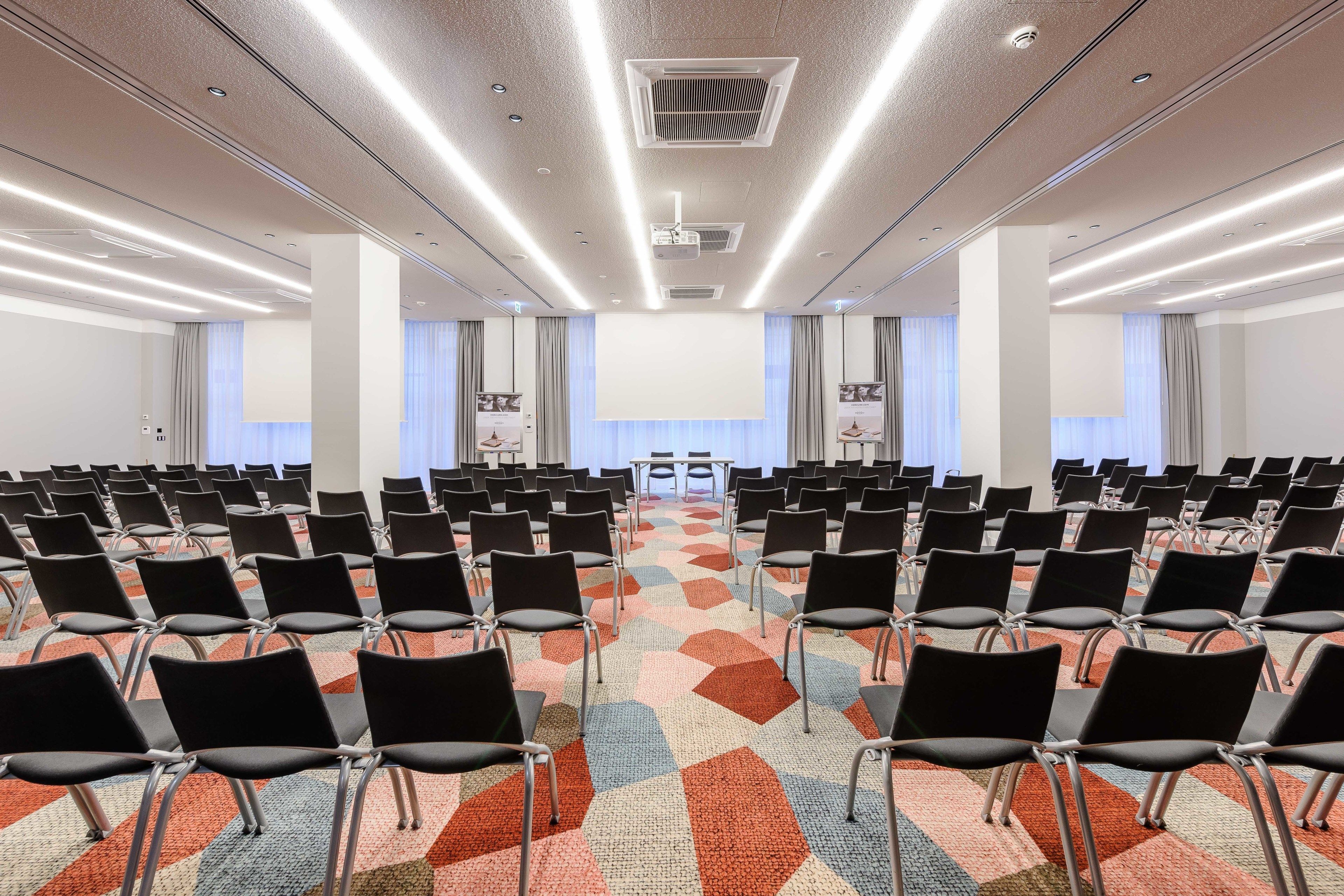 Uma sala de conferência com fileiras de cadeiras pretas, um tapete colorido e uma tela de projetor na frente.