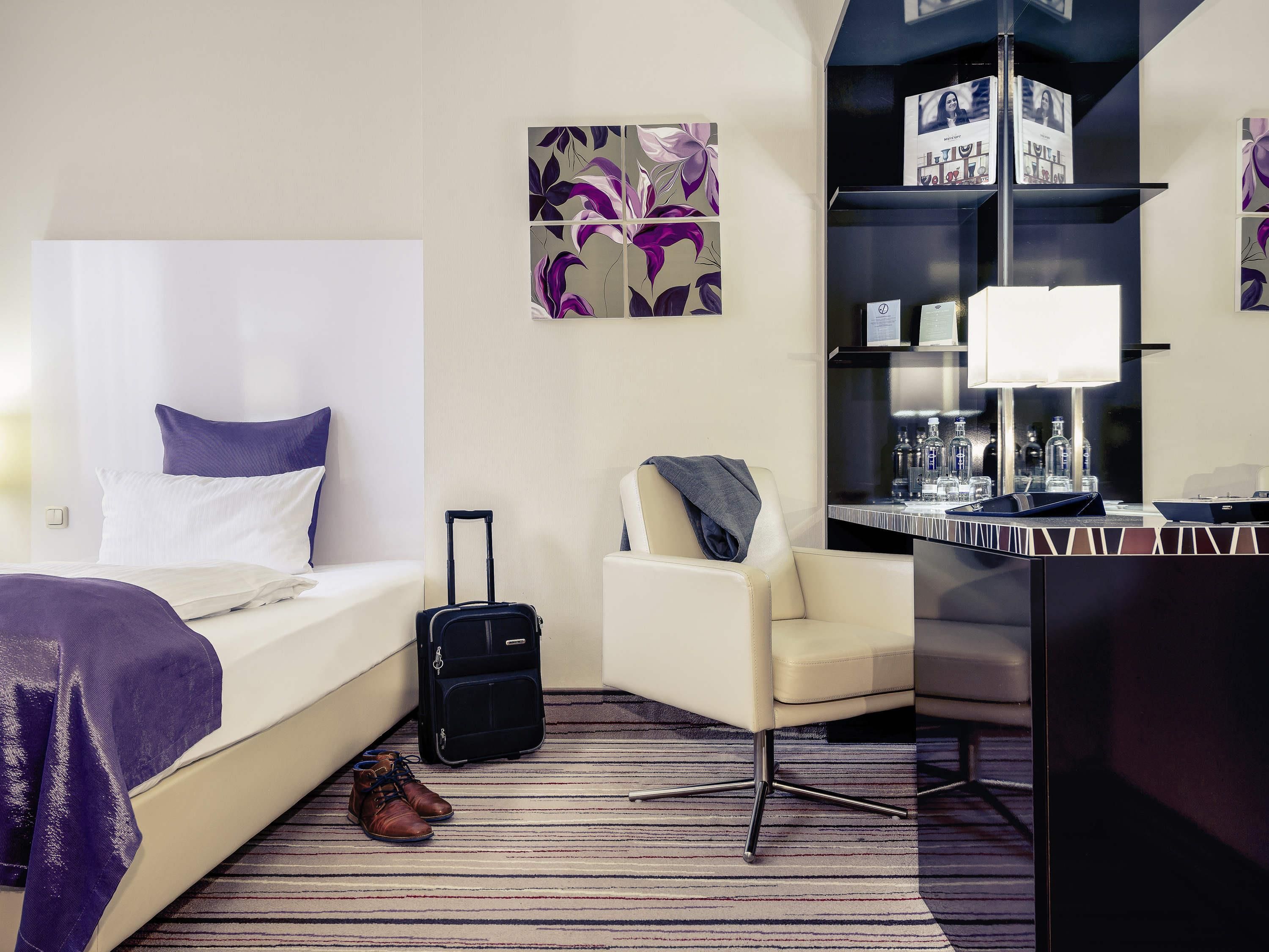 Quarto de hotel com cama, cadeira, escrivaninha, mala e minibar.
