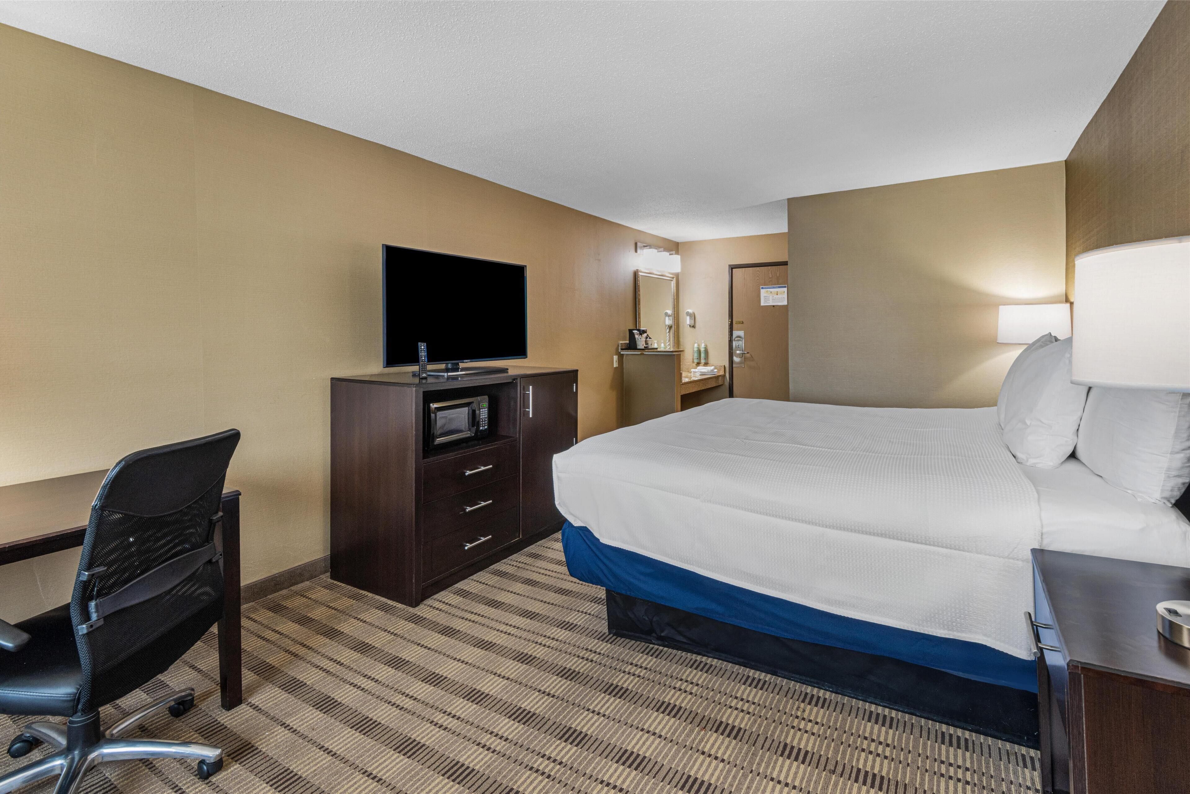 Quarto de hotel com cama, escrivaninha, cadeira, televisão e espelho.