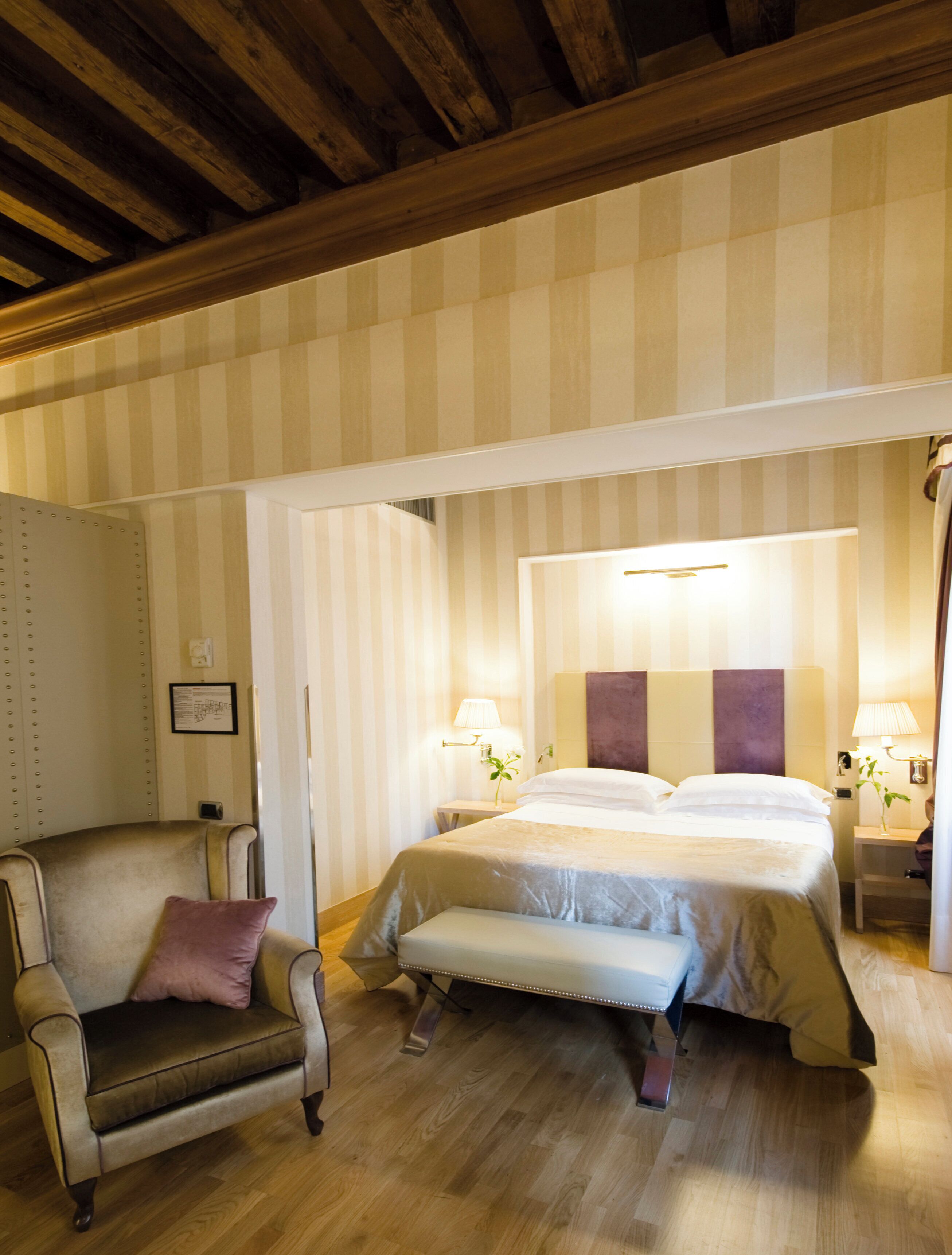 Quarto de hotel com cama, mesas de cabeceira, uma cadeira e papel de parede listrado.