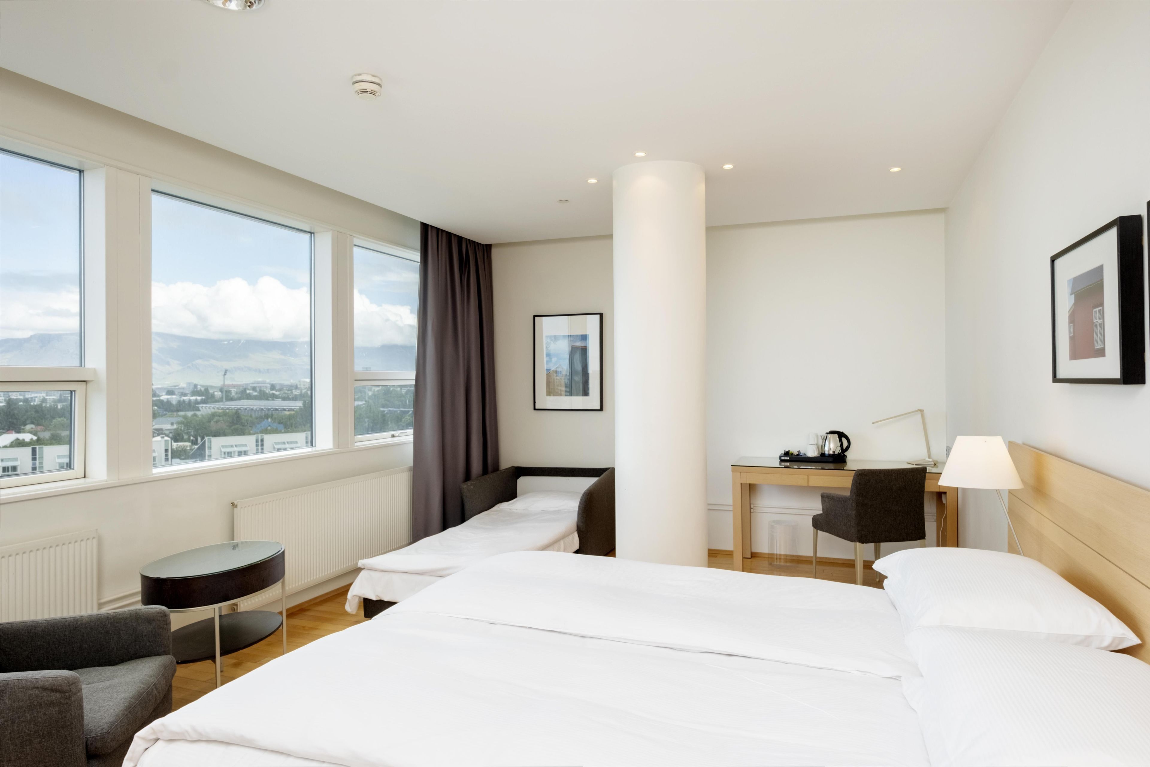 Quarto de hotel com duas camas, uma escrivaninha, uma cadeira, radiador e amplas janelas com vista para a cidade.