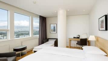 Quarto de hotel com duas camas, uma escrivaninha, uma cadeira, radiador e amplas janelas com vista para a cidade.