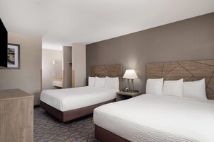 Quarto de hotel com duas camas, uma escrivaninha, um abajur e um espelho.
