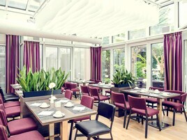 Uma zona de refeições moderna, com mesas preparadas para quatro pessoas, amplas janelas e plantas em vasos.