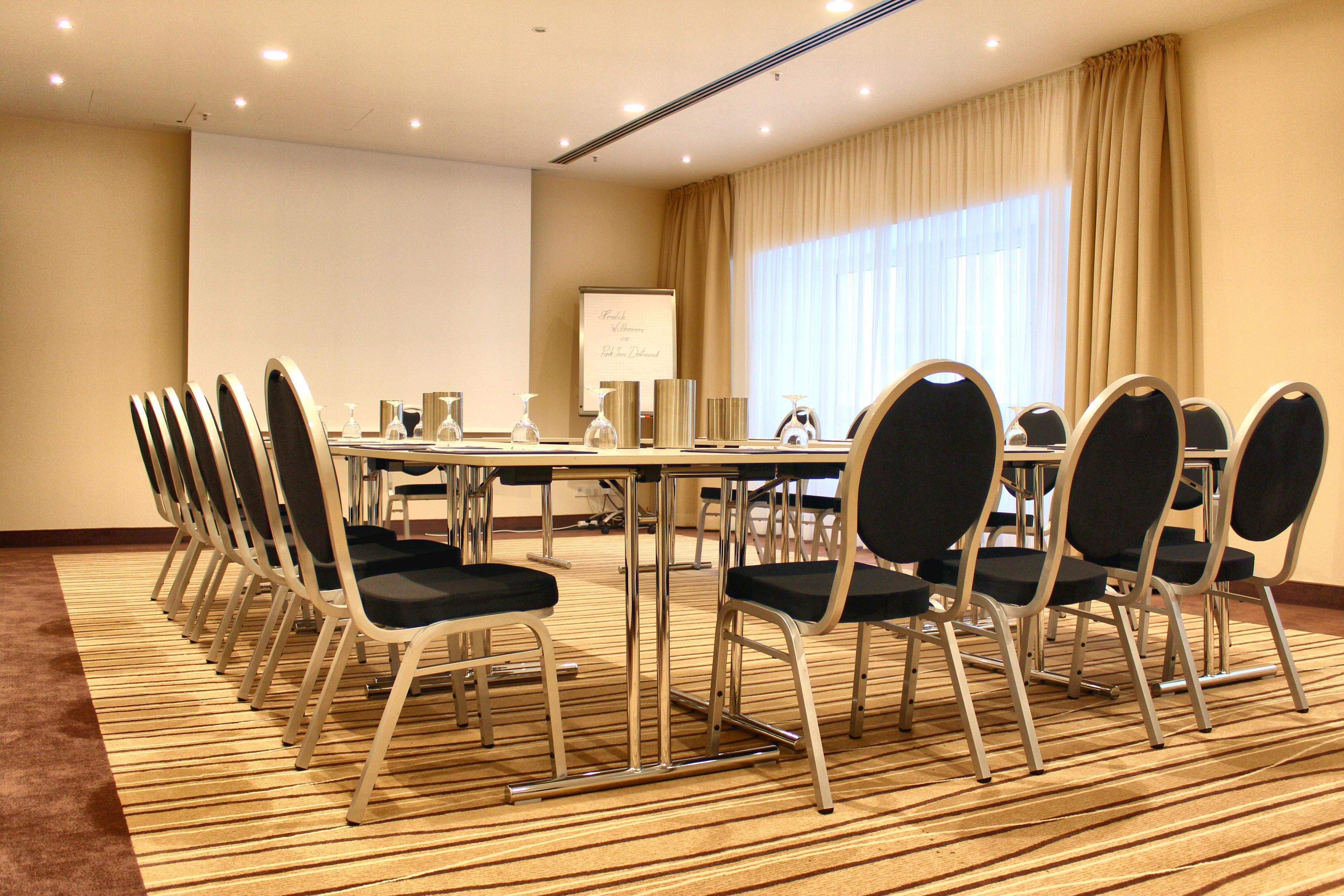 Uma sala de conferências com uma mesa comprida, cadeiras, um projetor e um grande janela com cortinas.