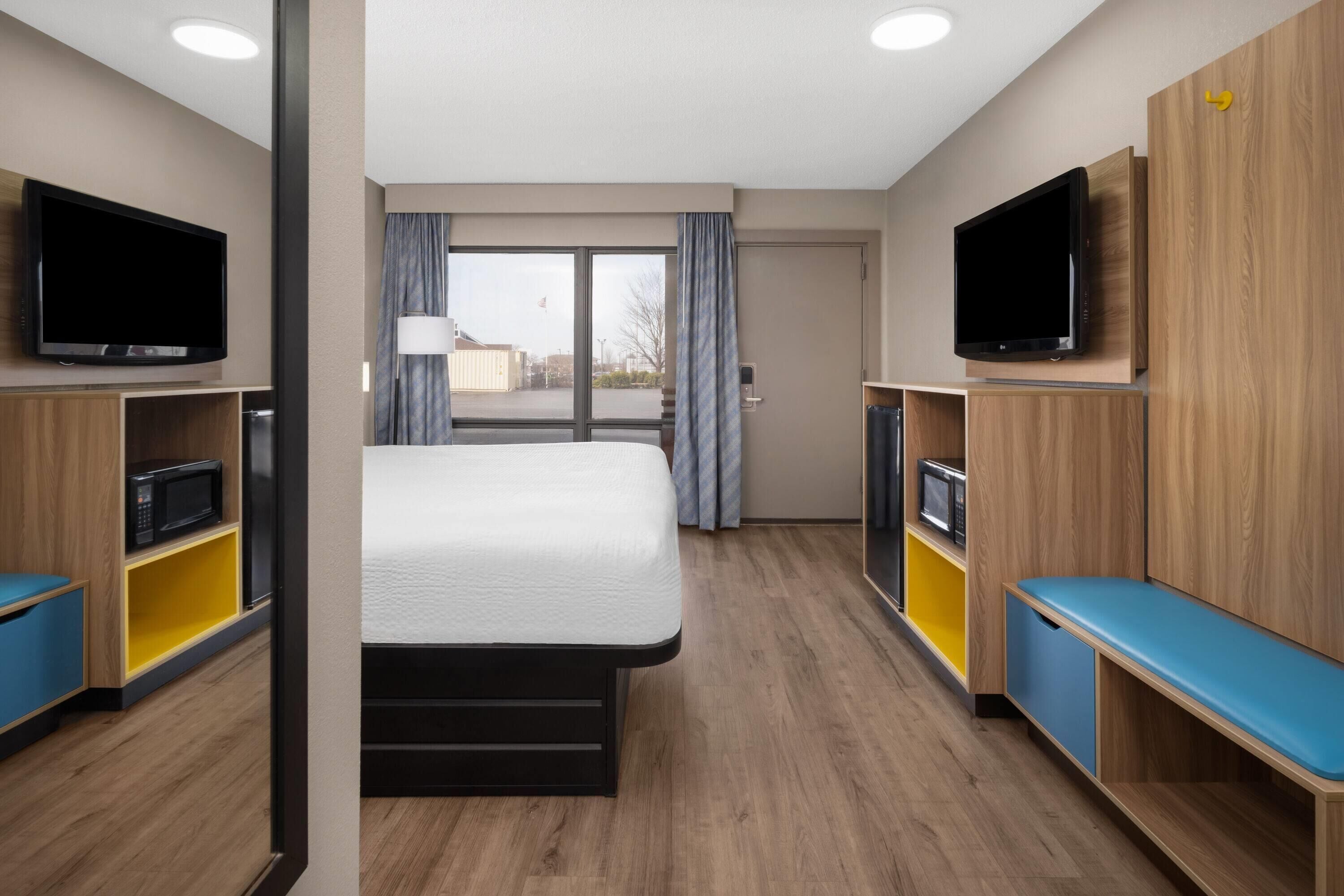 Quarto de hotel com cama, duas televisões, escrivaninha e vista para o exterior.