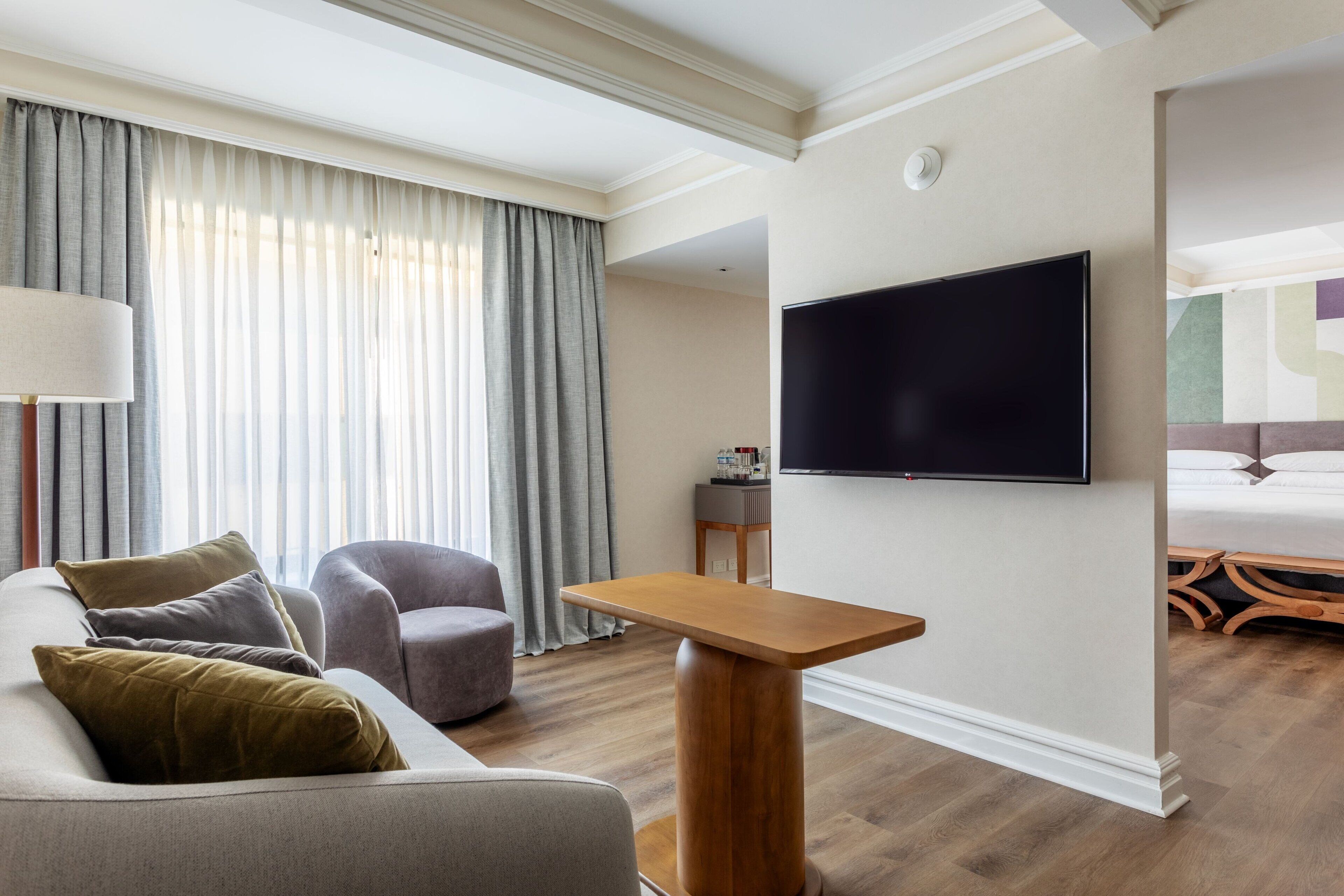 Quarto de hotel moderno com sofá, poltrona, televisão e piso de madeira.