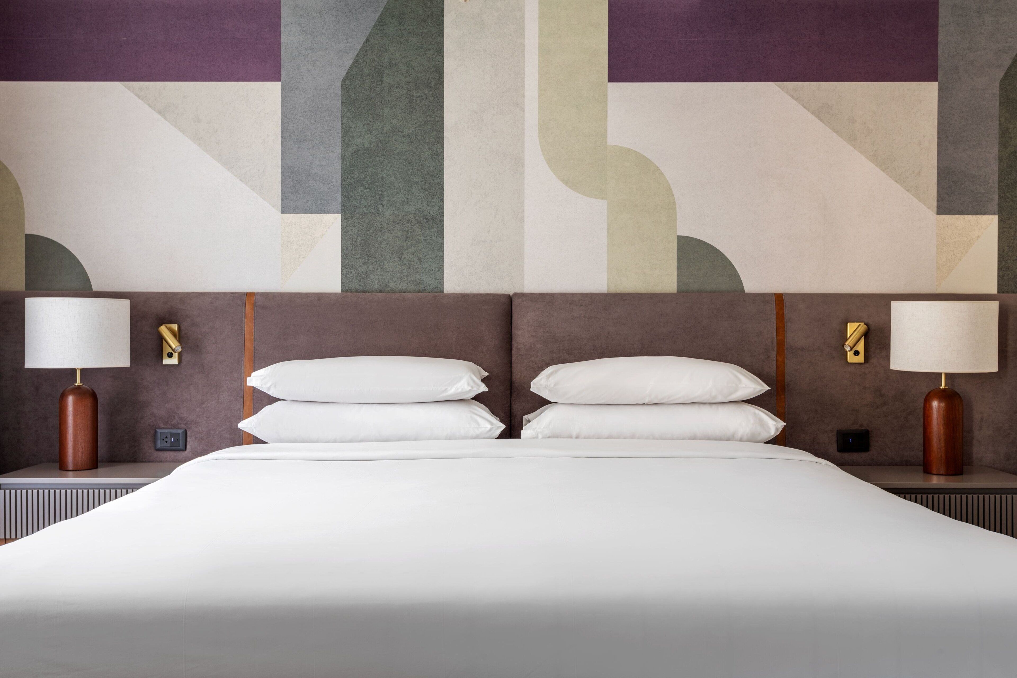 Quarto de hotel com uma cama grande, duas mesas de cabeceira com luminárias e uma parede com padrão geométrico.