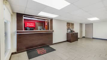 Uma área de recepção de hotel com placa vermelha indicando Wi-Fi gratuito em todos os quartos, um balcão de atendimento, uma máquina de café e uma porta para o Red Roof Inn.