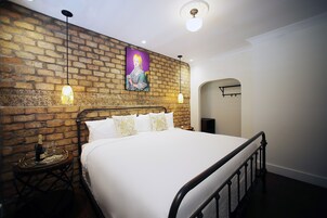Quarto de hotel com parede de tijolos, cama com roupa de cama branca, uma pequena mesa com uma garrafa e copos, e um quadro acima da cama.