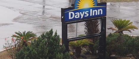 Placa do Days Inn com um logotipo de sol, cercada por vegetação e uma estrada molhada.