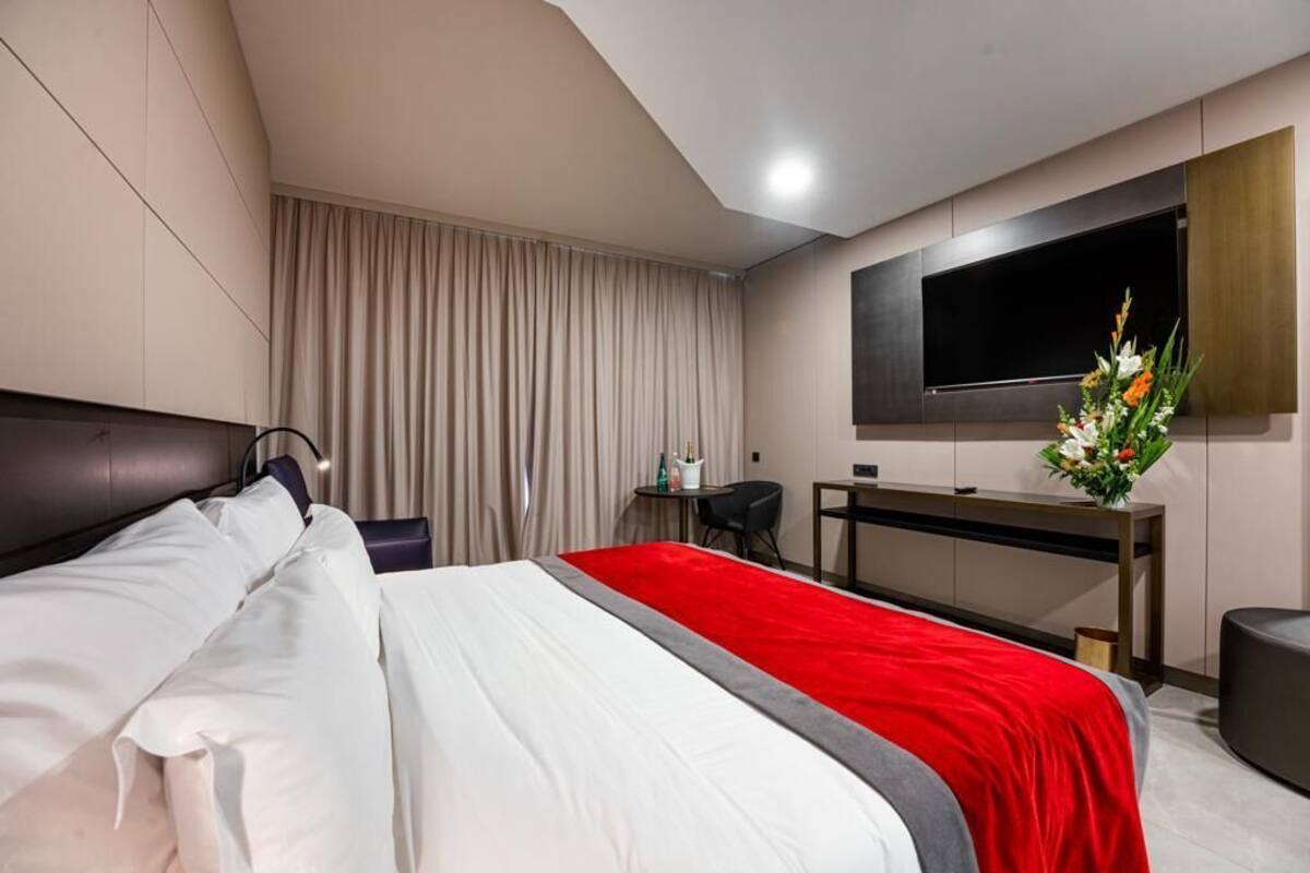 Quarto de hotel moderno com uma cama grande, uma escrivaninha, televisão de tela plana e uma janela ampla com cortinas.