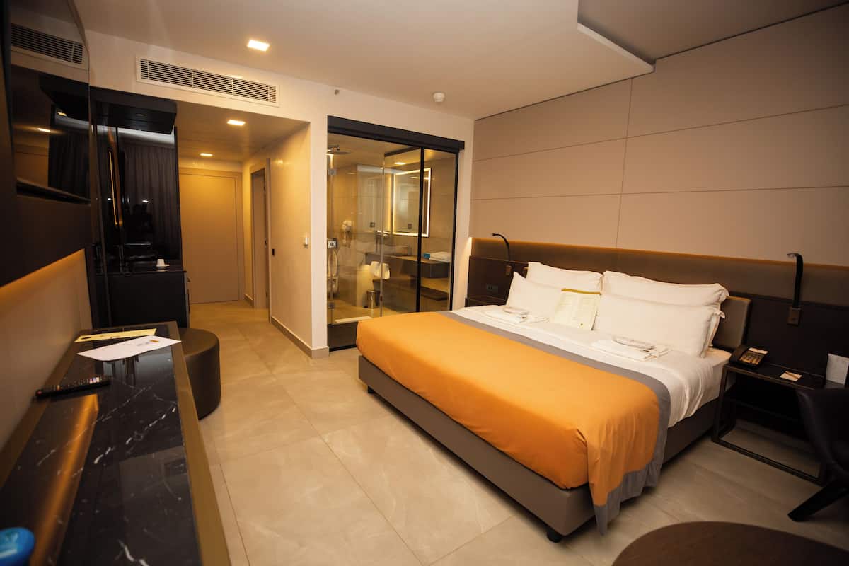 Quarto de hotel com cama, escrivaninha, cadeira, espelho e banheiro visível por uma porta aberta.