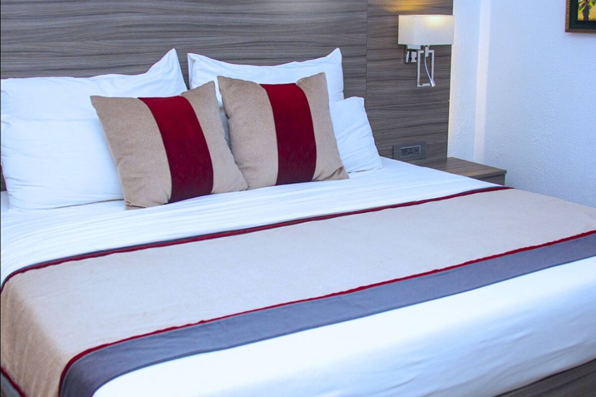 Cama bem arrumada com colcha branca e vermelha, duas almofadas com desenho vermelho e bege, e cabeceira de madeira.