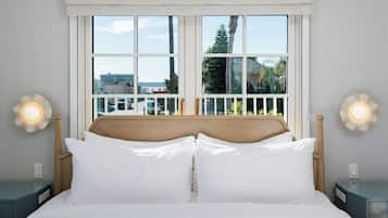 Cama bem arrumada com lençóis brancos, cabeceira de madeira e vista pela janela grande para edifícios e árvores.