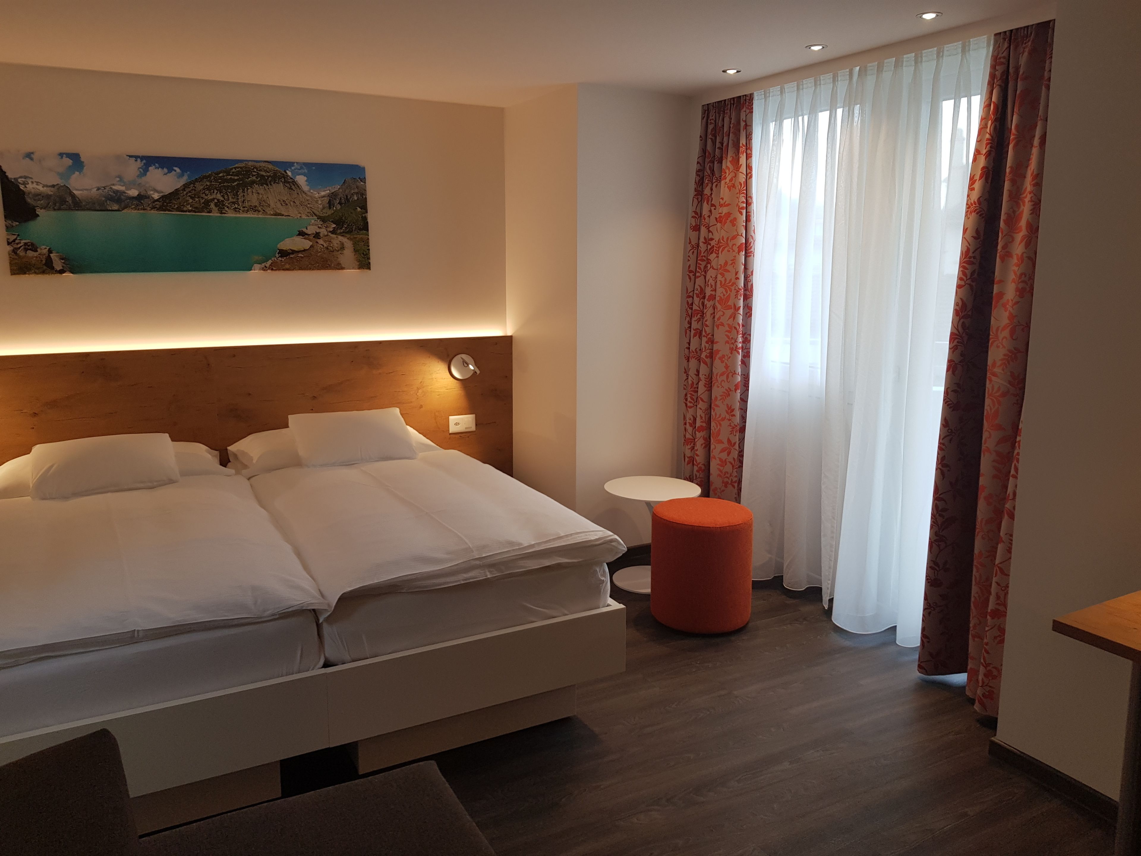 Quarto de hotel com cabeceira de madeira, cama com roupa branca, um banquinho redondo laranja, uma mesa redonda e uma pintura de uma paisagem montanhosa.