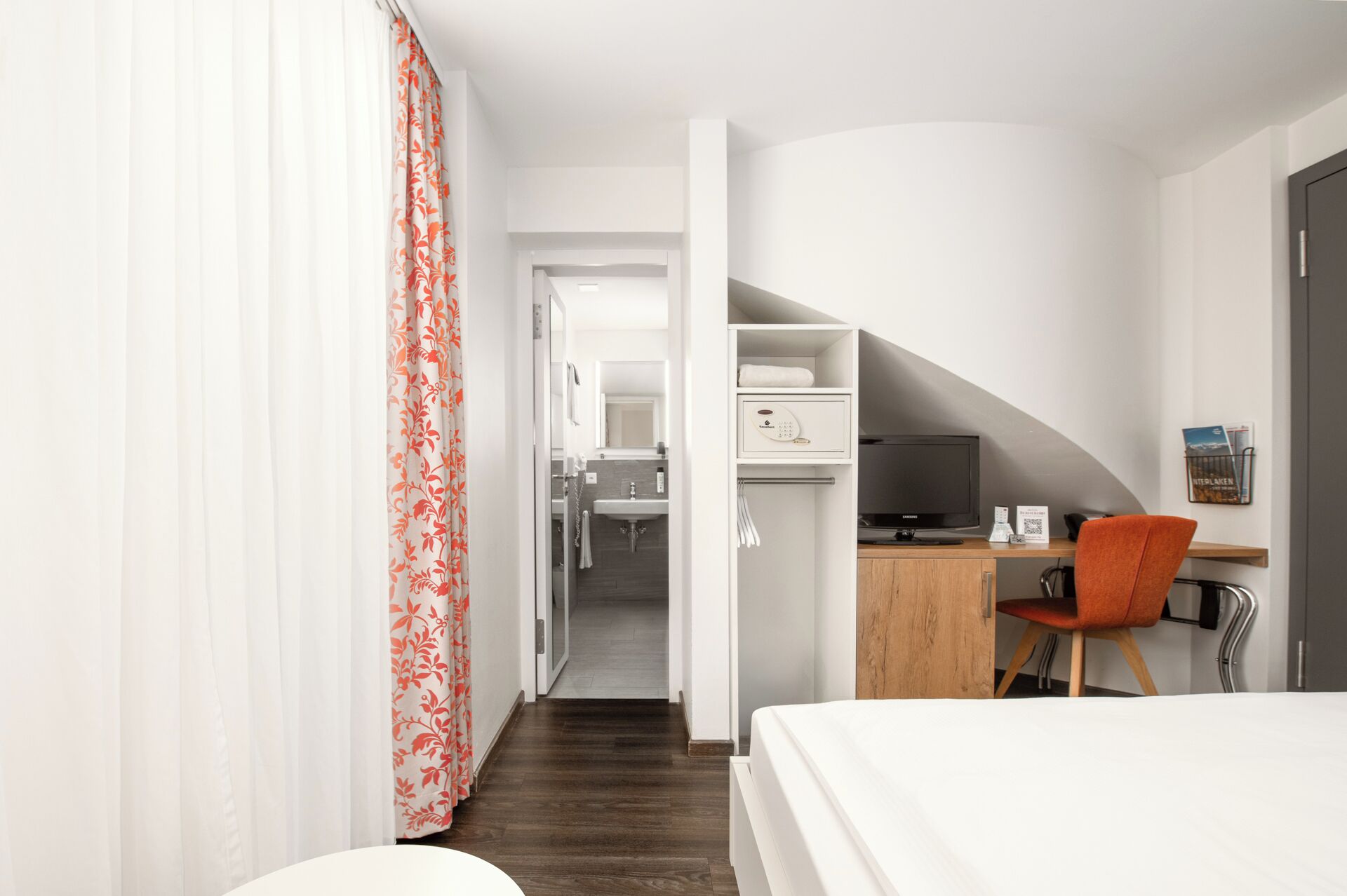 Quarto de hotel com uma cama, uma escrivaninha com computador, uma cadeira, banheiro com espelho e chuveiro.