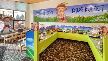 Espaço de buffet infantil com um mural de uma criança e elementos da natureza, mesas com comida e um tapete estampado.