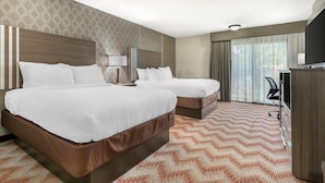 Quarto de hotel com duas camas, uma escrivaninha, uma luminária e uma janela grande com cortinas.