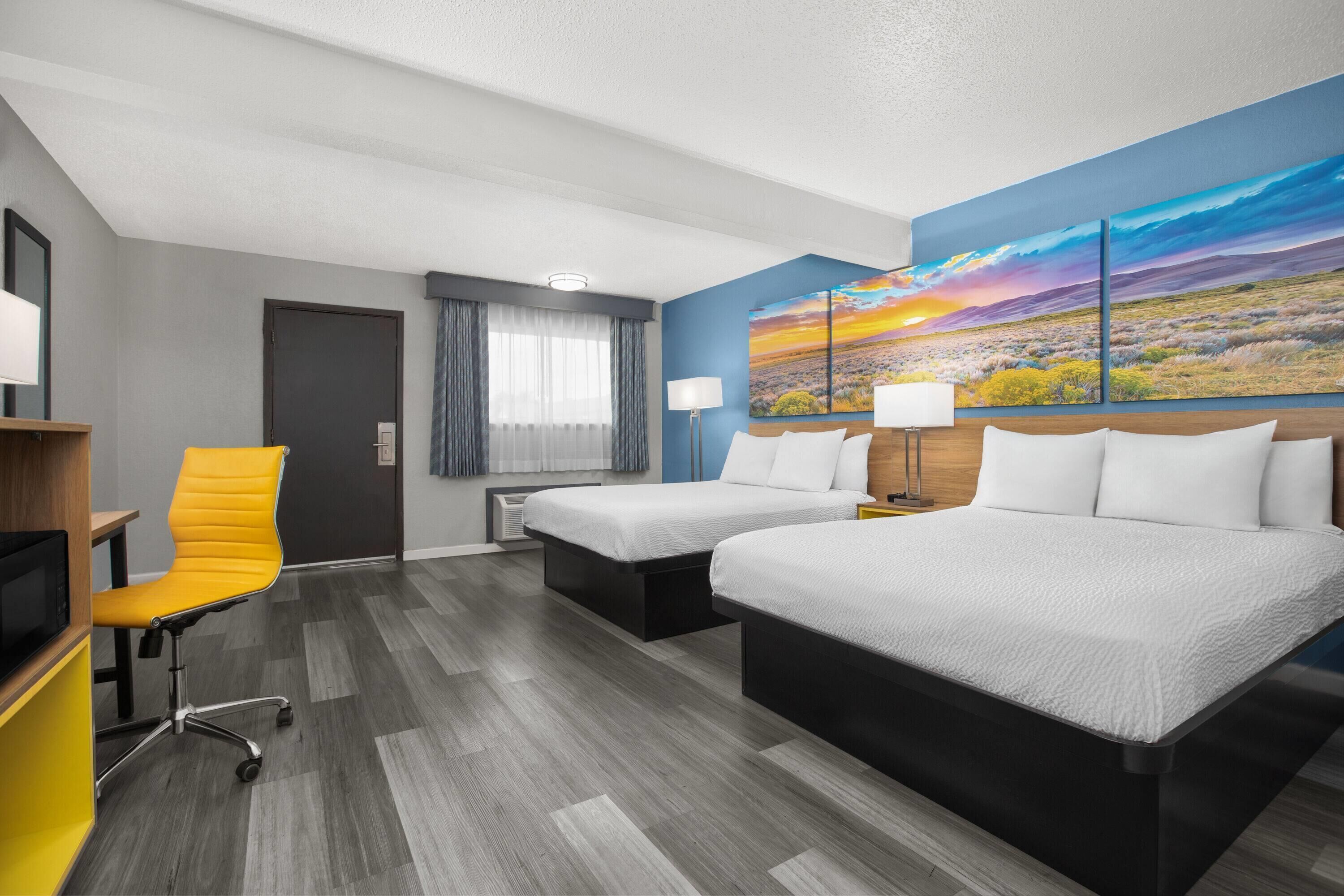 Quarto de hotel com duas camas, uma escrivaninha, uma cadeira, televisão e uma grande pintura de paisagem na parede.