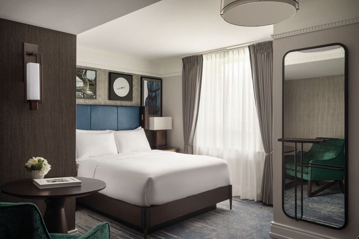 Quarto de hotel com uma cama grande, uma mesa redonda com um vaso de flores, uma cadeira verde, um espelho e vista da cidade pela janela.