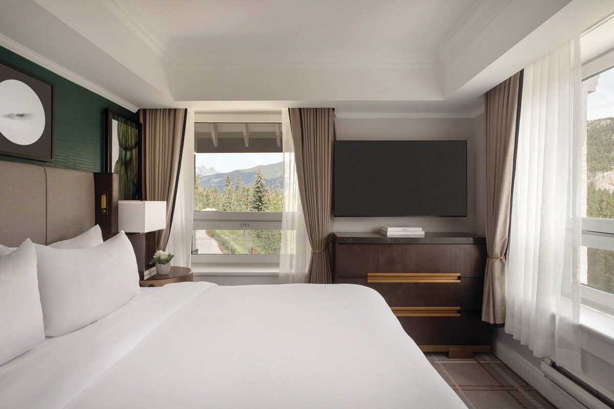 Quarto de hotel com uma cama grande, uma escrivaninha com televisão de tela plana, uma janela com cortinas e vista para montanhas.