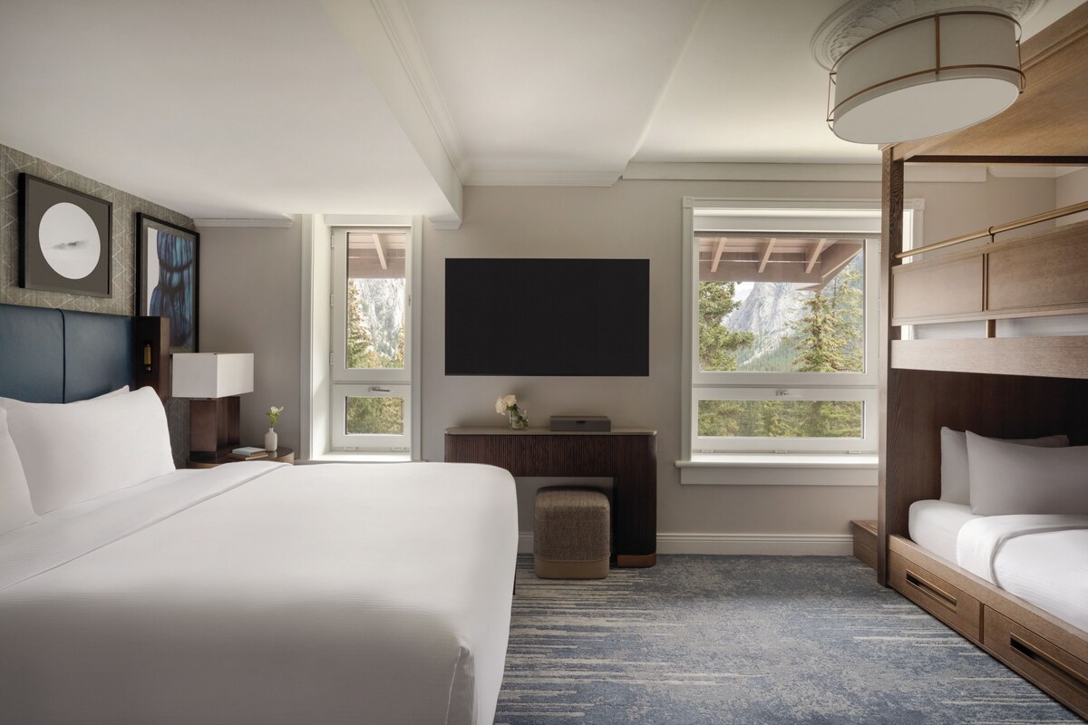 Quarto de hotel com uma cama grande, uma escrivaninha com televisão de tela plana, uma janela com vista para árvores e uma cama beliche.