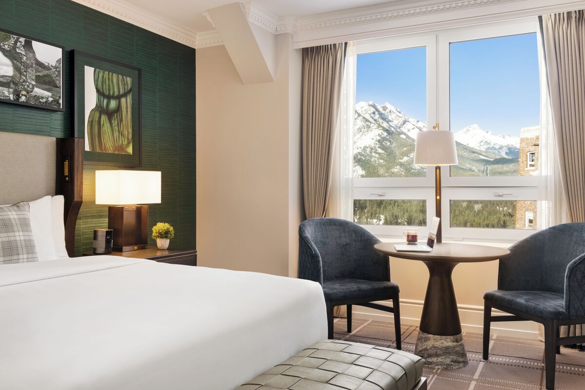 Quarto de hotel com uma cama grande, duas poltronas, uma mesinha, um abajur e vista para as montanhas pela janela.