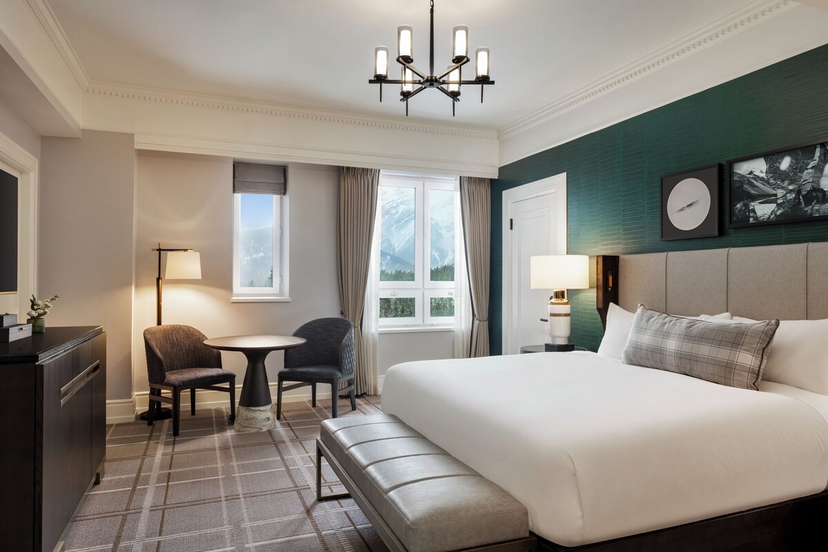 Quarto de hotel com uma cama grande, uma escrivaninha, uma cadeira, um banco, um lustre, uma janela com vista para montanhas e uma porta.