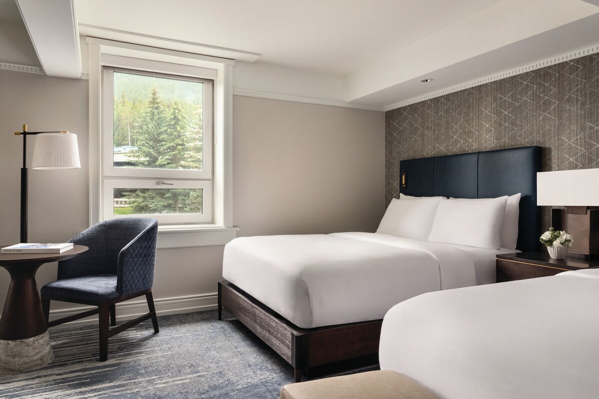 Quarto de hotel com uma cama grande, uma cadeira, uma mesinha e uma janela com vista para árvores.