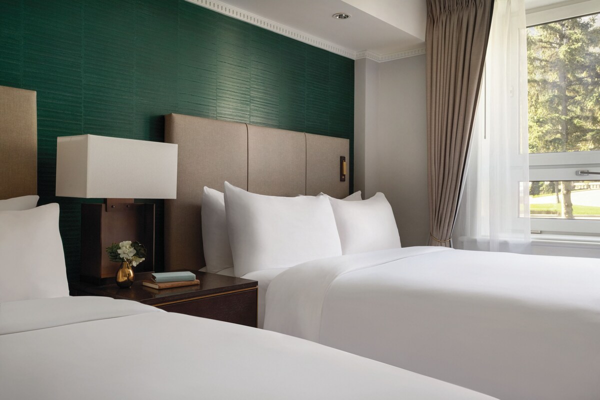 Quarto de hotel com duas camas, uma mesa de cabeceira, uma luminária e uma janela com cortinas.