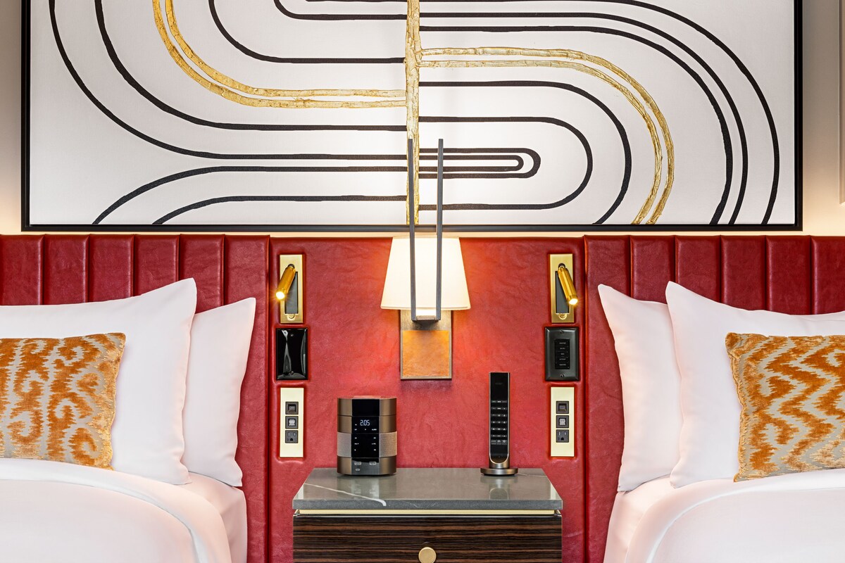 Quarto de hotel com design moderno, apresentando uma grande obra de arte abstrata na parede, um painel de cabeceira estofado em vermelho, uma mesa de cabeceira com abajur e duas almofadas com estampas.