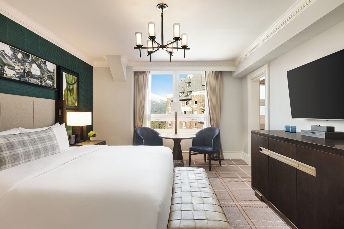 Quarto de hotel com uma cama grande, uma mesa de trabalho com televisão de tela plana, um candelabro e vista para prédios pela janela.