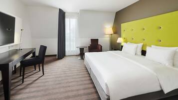 Quarto de hotel com uma cama grande, uma escrivaninha, uma cadeira, uma televisão e um abajur.