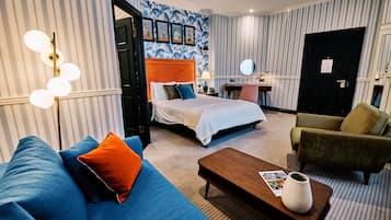 Quarto de hotel com uma cama, um sofá azul, uma mesa de centro de madeira, uma poltrona verde e uma parede listrada com padrão tropical.