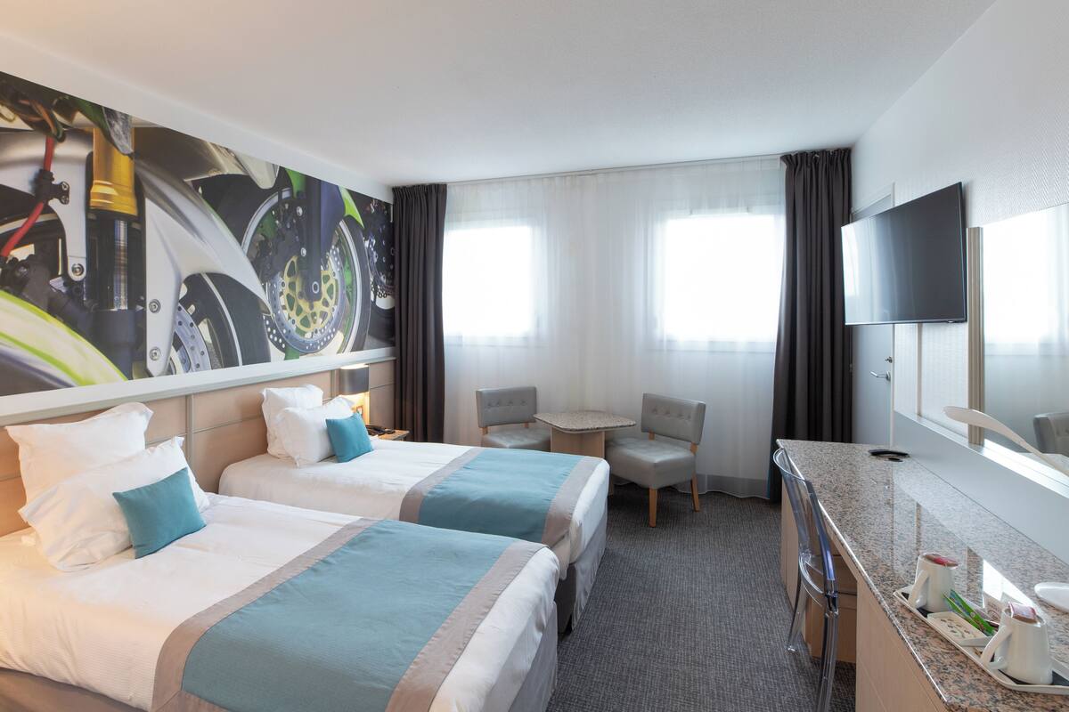 Quarto de hotel com duas camas, uma mesa, uma cadeira, uma televisão e uma janela grande com cortinas.