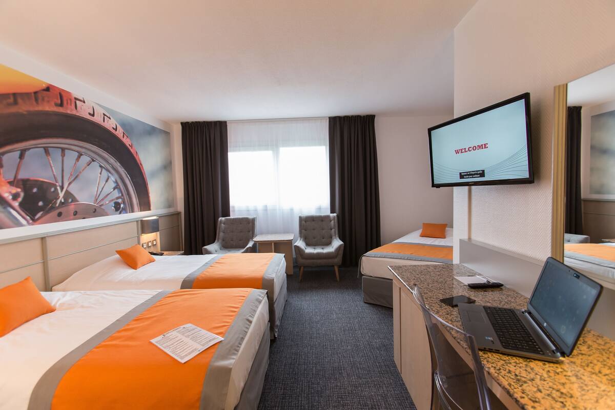 Quarto de hotel com duas camas, uma secretária com um laptop, uma TV a exibir "WELCOME" e uma janela grande com cortinas.