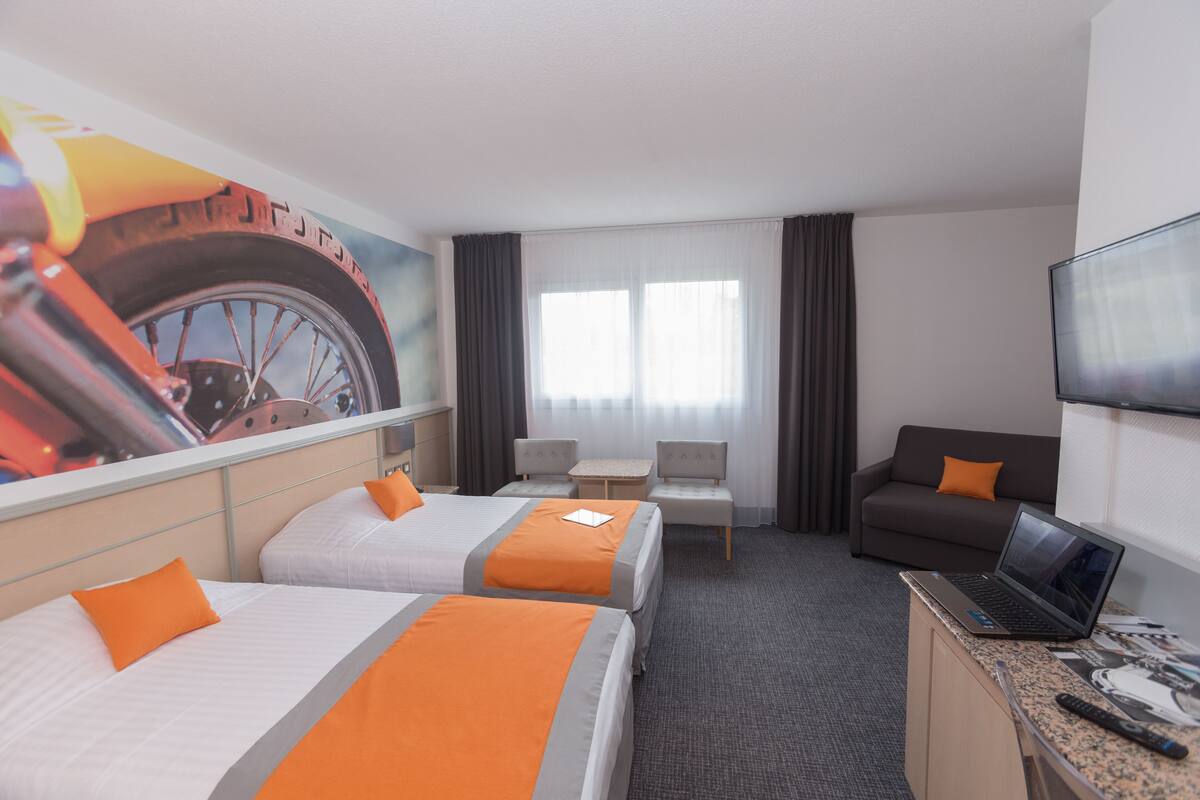 Quarto de hotel com duas camas, uma mesa com um laptop, uma televisão e uma janela grande com cortinas.