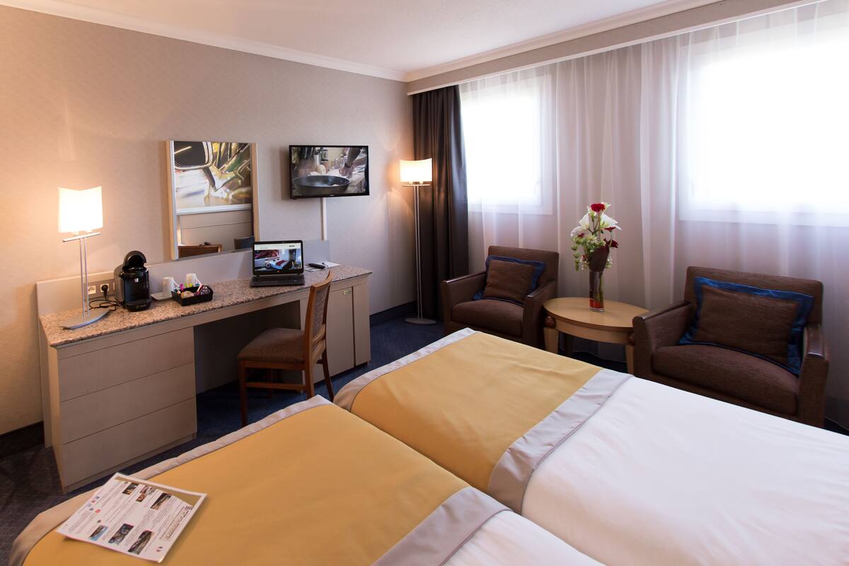 Quarto de hotel com cama, escrivaninha, cadeiras, televisão e um vaso com flores.