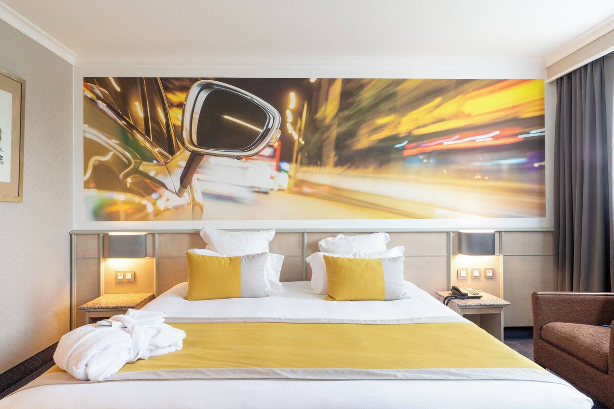 Quarto de hotel com uma cama grande, duas mesas de cabeceira com abajures, uma cadeira e um quadro abstrato grande na parede.