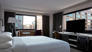 Quarto de hotel com uma cama grande, uma escrivaninha com televisão, uma cadeira e vista para edifícios pela janela.