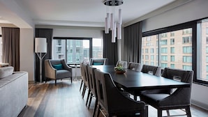 Quarto de hotel moderno com uma grande mesa de jantar, assentos confortáveis, abajur e janelas amplas com vista para a cidade.