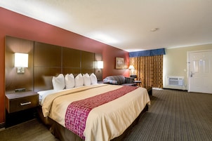 Quarto de hotel com uma cama grande, mesa de cabeceira, escrivaninha, cadeira, abajur, quadro na parede e uma porta.