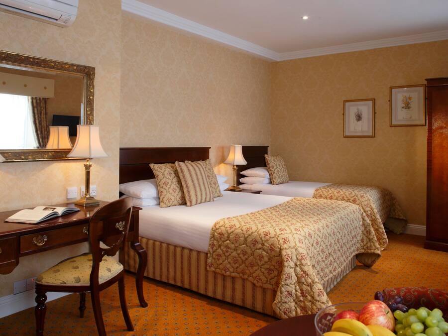 Quarto de hotel com duas camas, uma escrivaninha com luminária, uma cadeira e uma fruteira no chão.