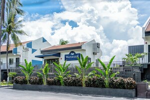 Um hotel chamado Waterside Suites & Marina, com design arquitetônico moderno, apresenta uma placa bem visível, um jardim bem cuidado com plantas tropicais e um céu azul claro com nuvens.